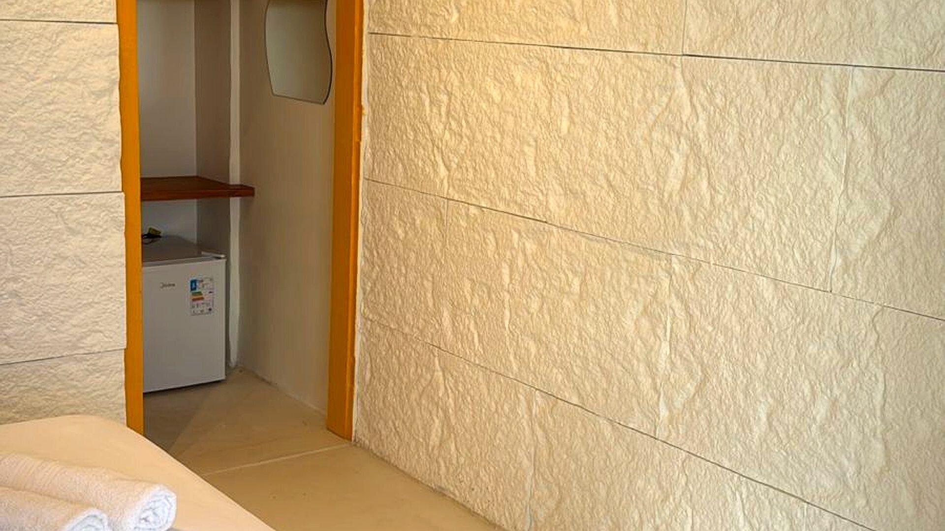 Um corredor estreito com parede branca texturizada, moldura de porta de madeira e uma pequena geladeira ao fundo.