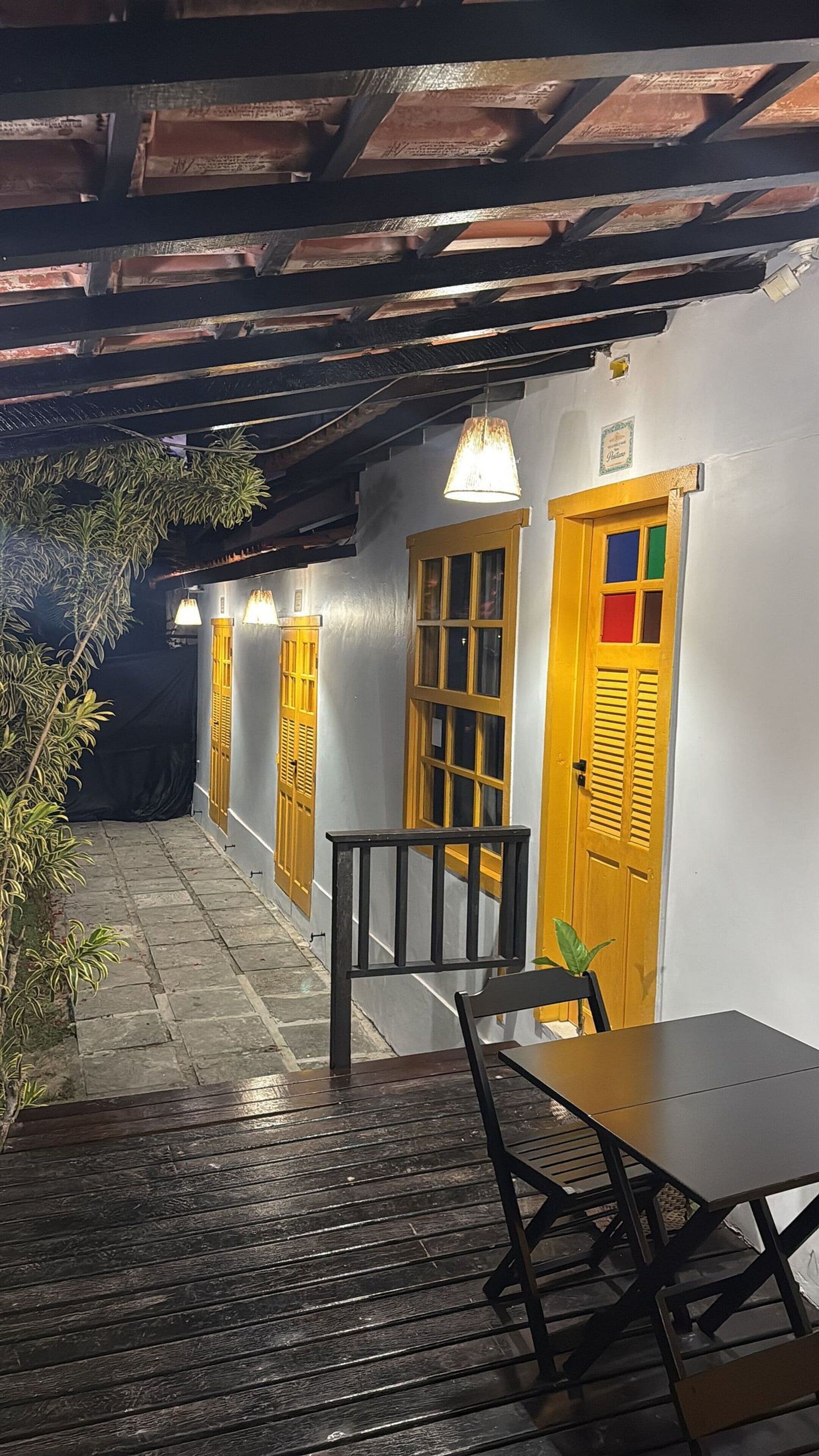 Uma área de estar ao ar livre coberta, com mesa, cadeiras, portas de madeira e um lustre pendurado.