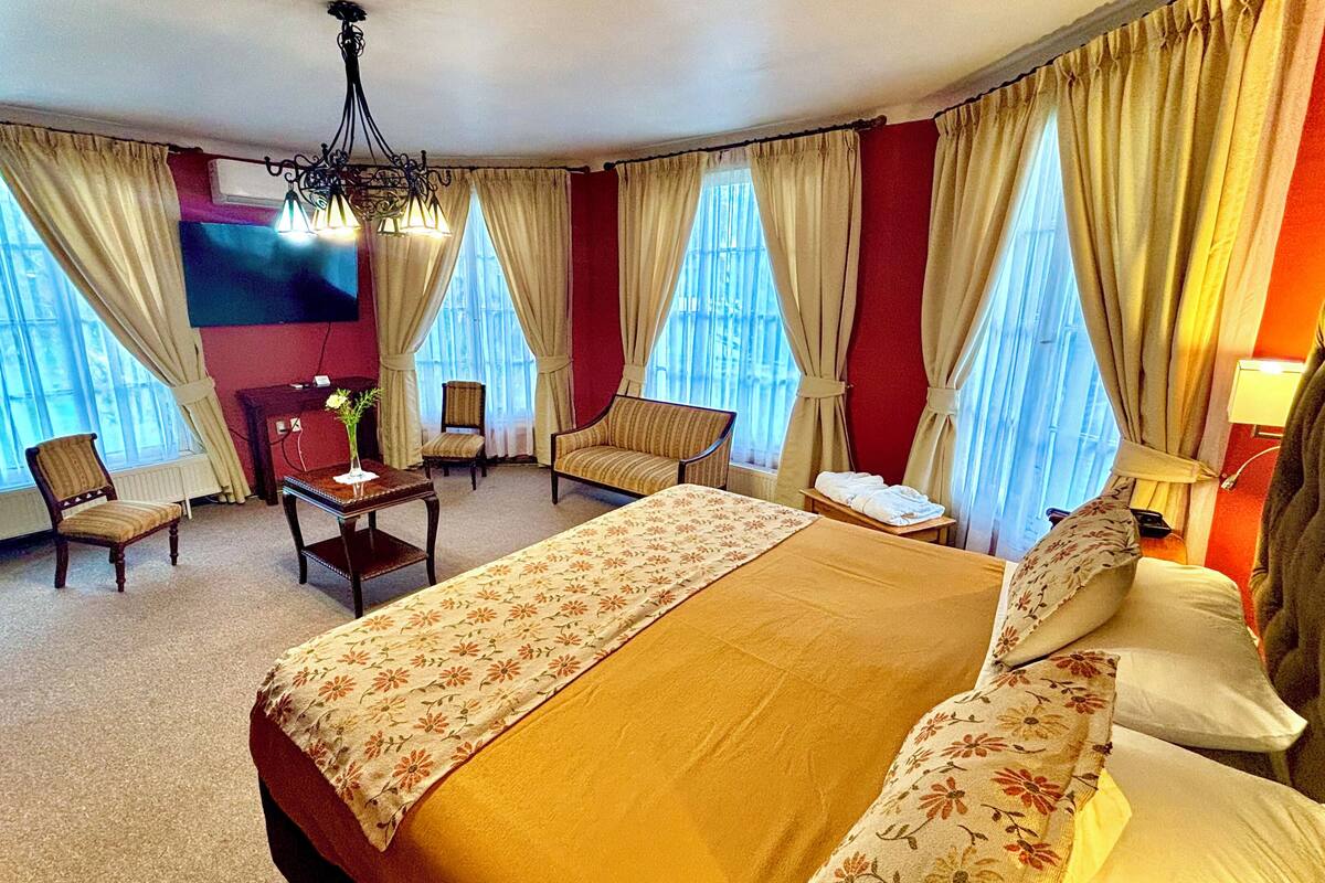 Quarto de hotel com uma cama grande, duas cadeiras, uma mesinha de centro com um vaso e uma televisão na parede.