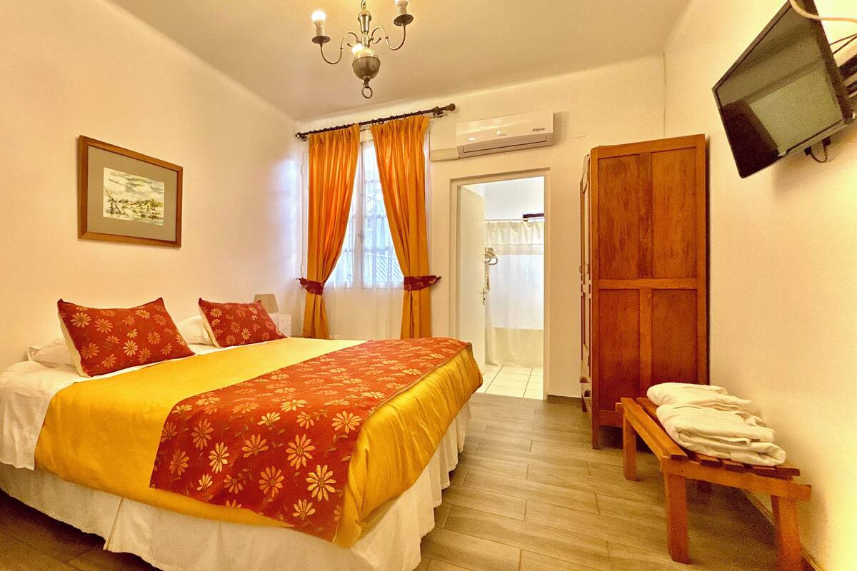 Quarto de hotel com uma cama, televisão, um banco de madeira e uma porta que dá para o banheiro.