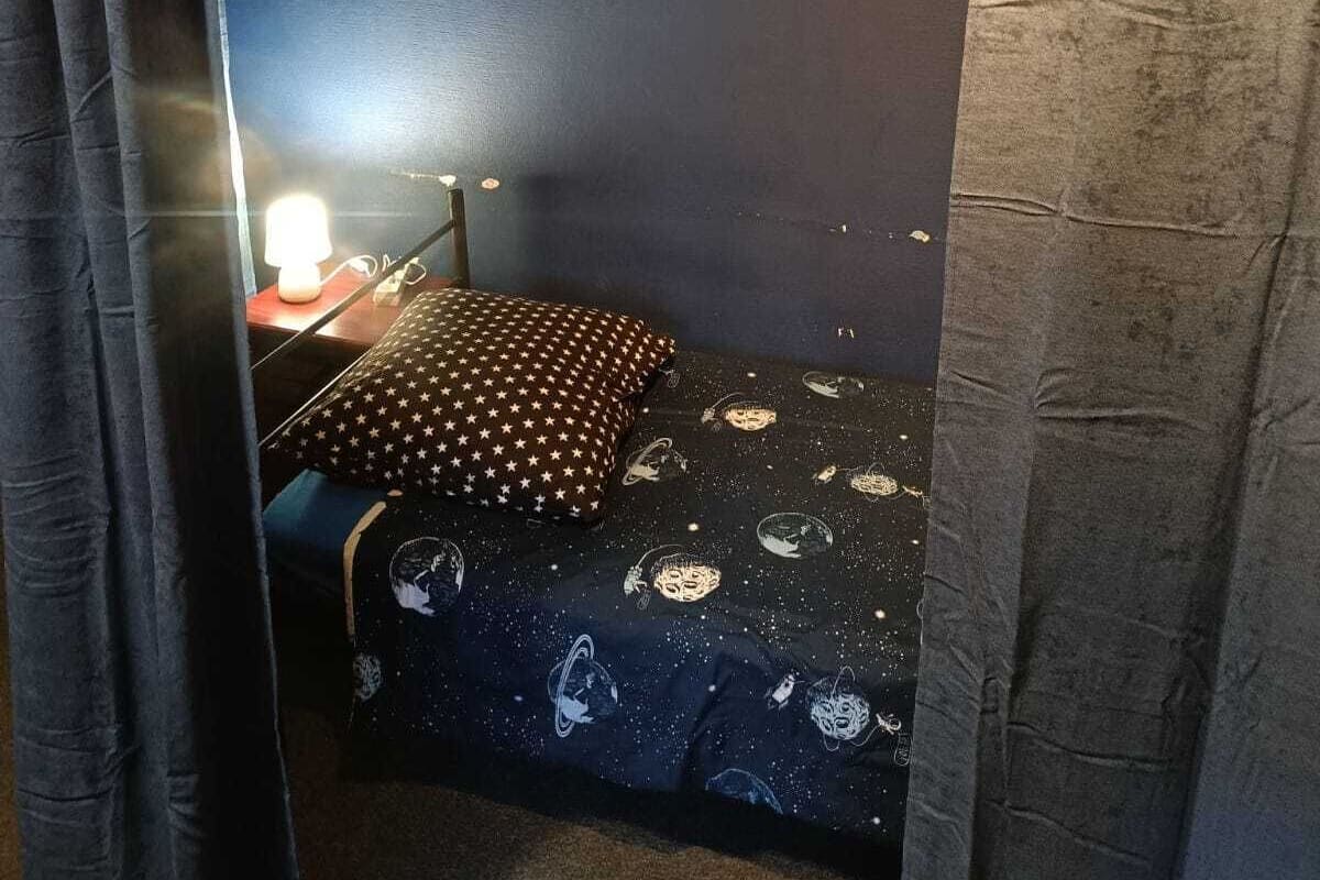 Quarto pequeno com uma cama de solteiro, parede azul escura, um número '1' indicando a cama, mesa de cabeceira com abajur e uma janela com cortinas.