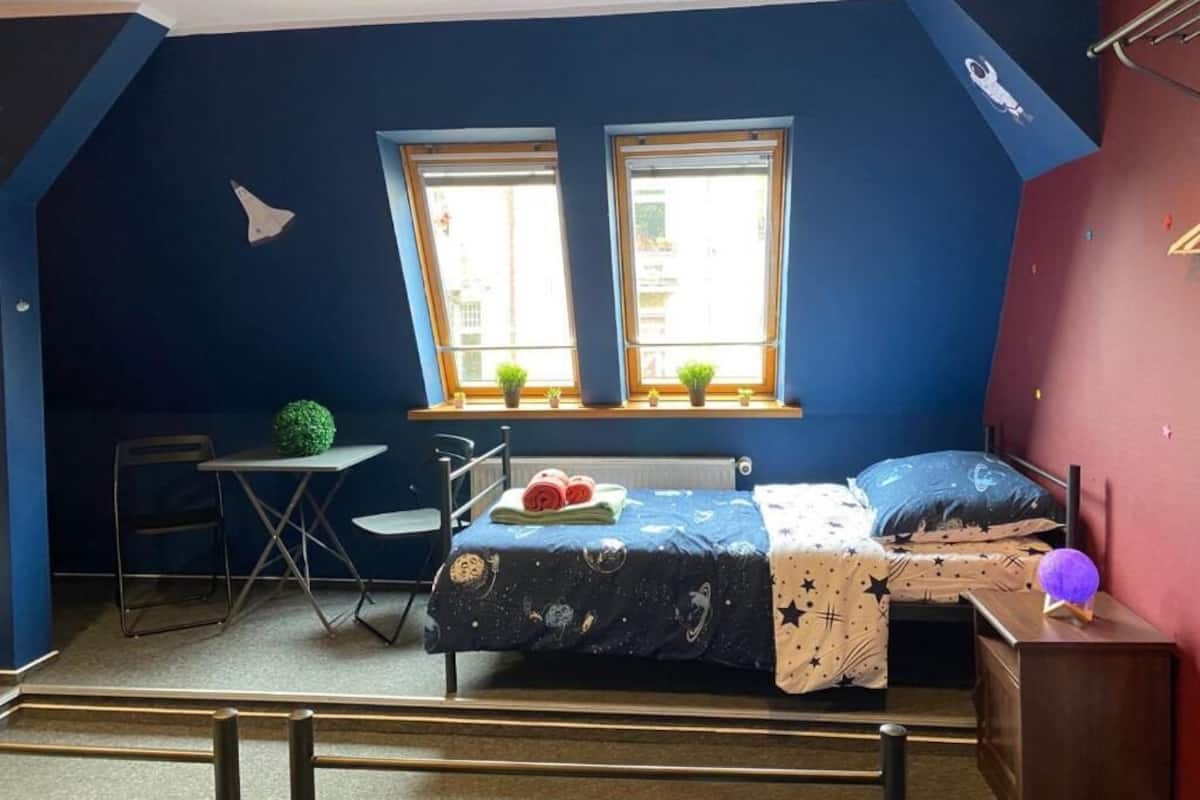 Quarto com cama, duas janelas, uma escrivaninha e uma cadeira.