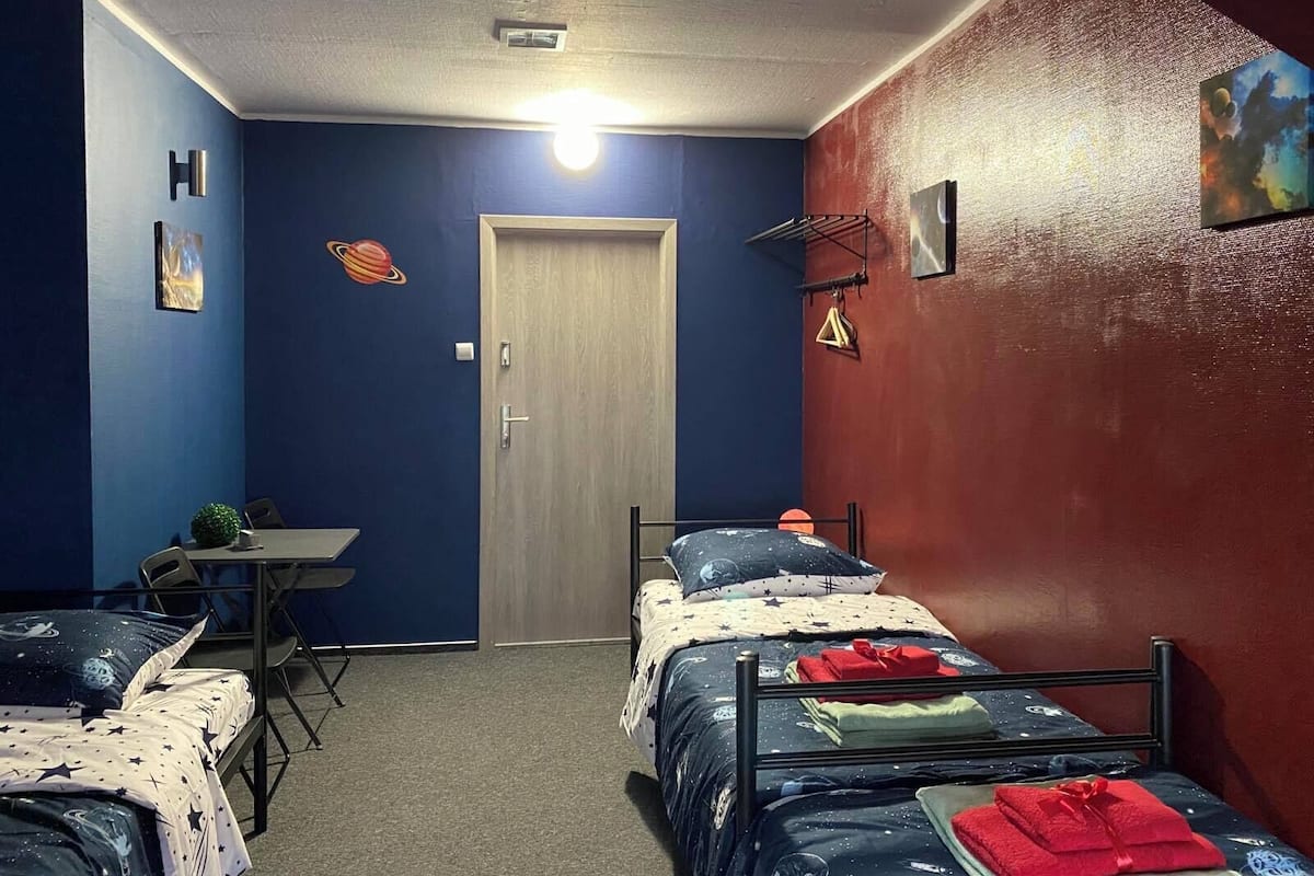 Quarto com duas camas, uma escrivaninha, uma cadeira, uma porta e uma parede com um quadro.