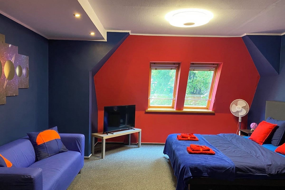 Quarto com televisão, duas camas, sofá, ventilador e duas janelas.