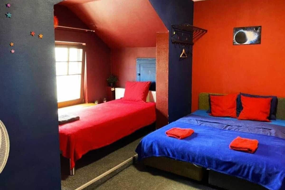 Quarto com duas camas, uma com roupa de cama vermelha e outra com roupa de cama azul, uma janela e um ventilador fixado na parede.