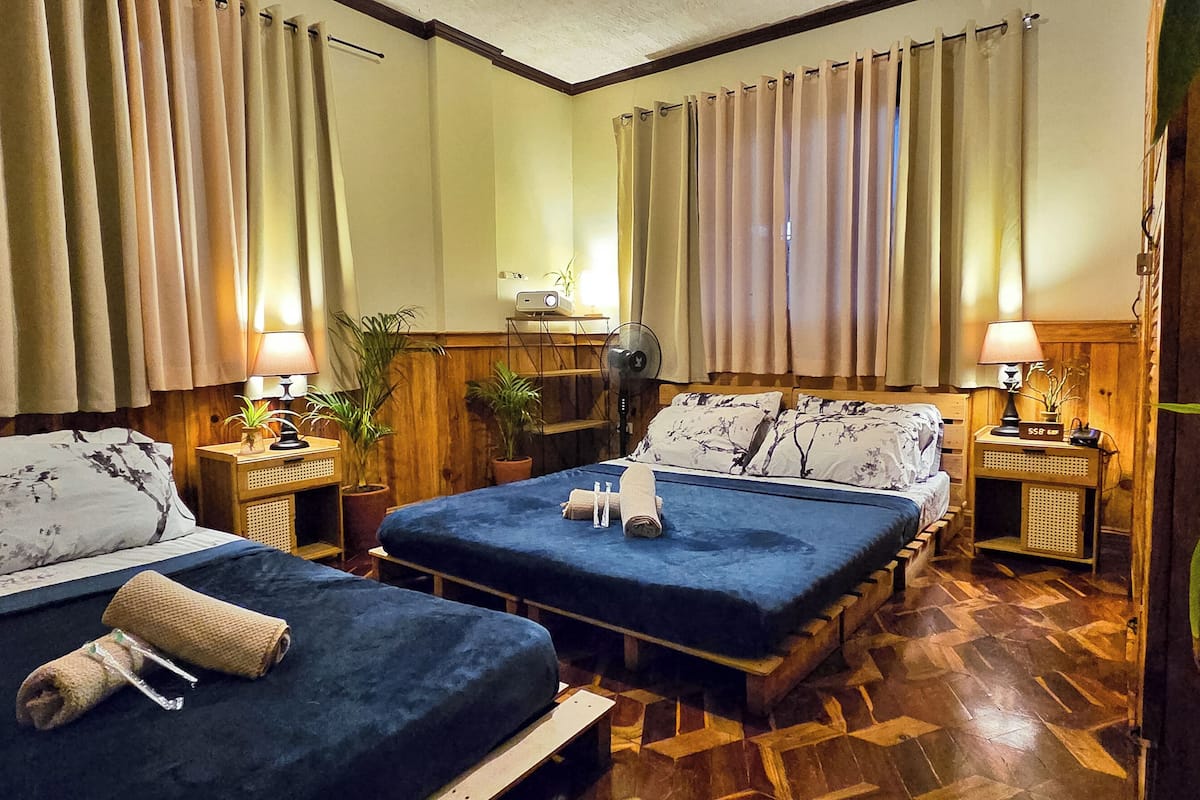 Quarto de hotel com duas camas, piso de madeira, cortinas, plantas em vasos e luminárias de cabeceira.