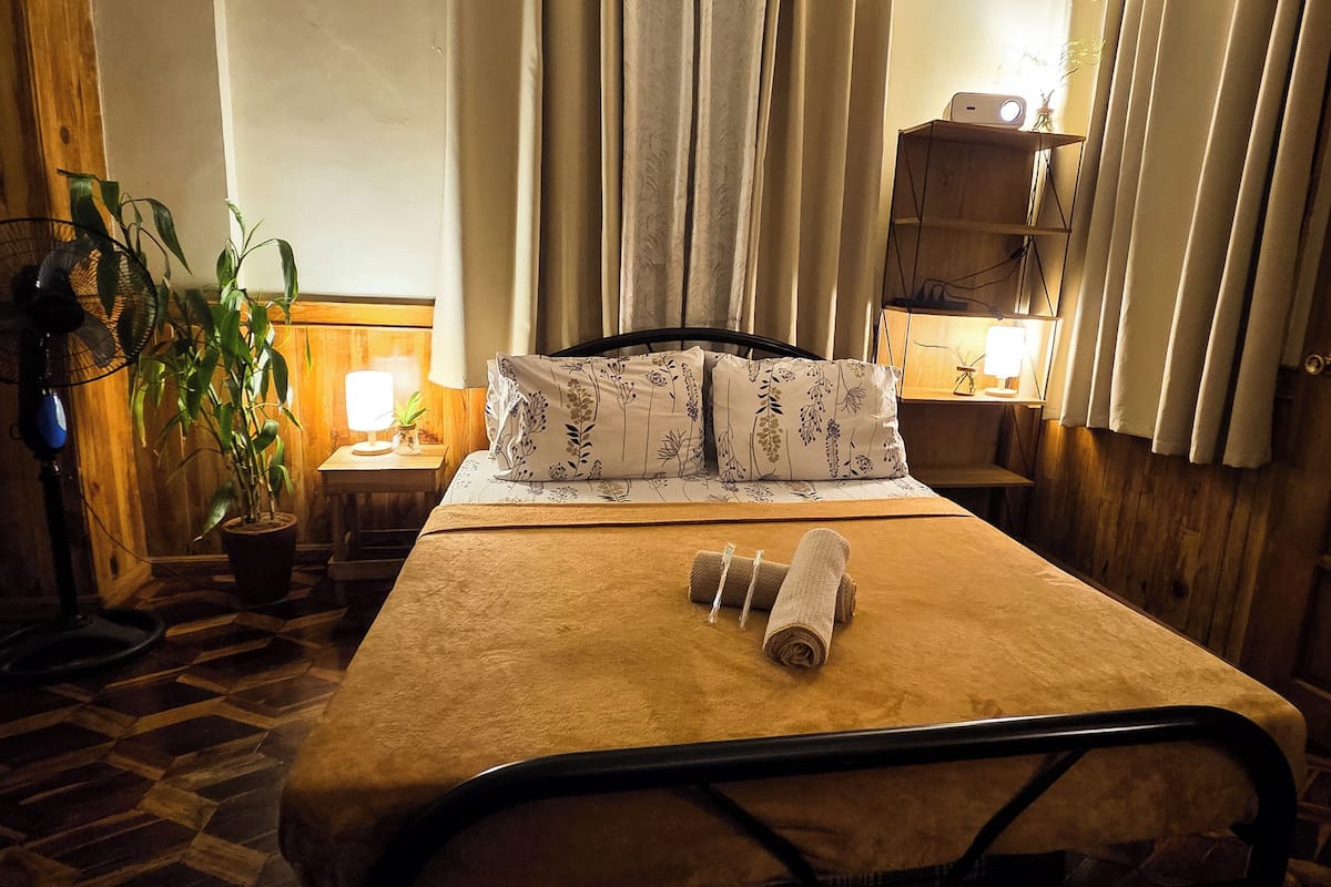 Um quarto com uma cama, uma mesa de cabeceira com abajur, um ventilador e uma estante com luminária.