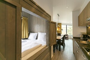 Quarto com cabeceira de madeira, cama com roupa de cama branca, área de jantar com mesa e cadeiras e cozinha com pia e fogão.