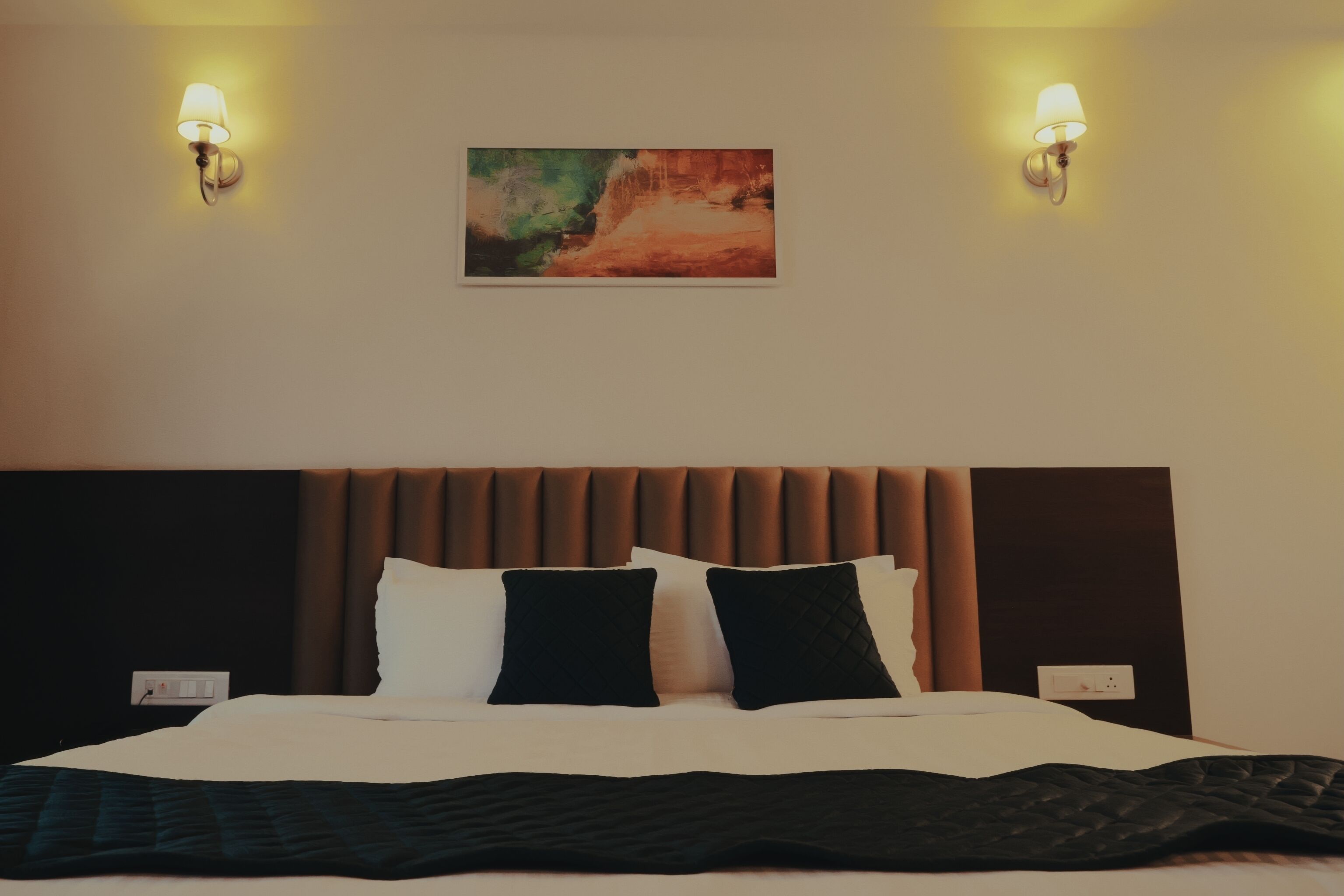 Quarto de hotel com uma cama, duas luminárias na parede e um quadro acima da cama.