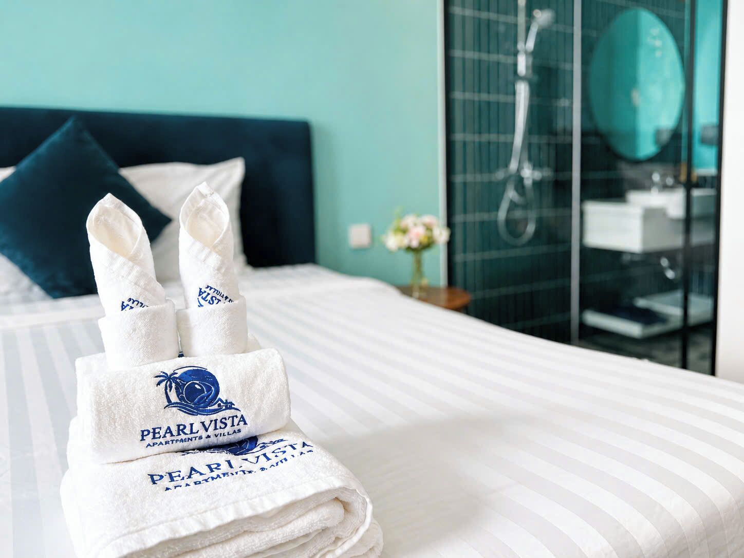 Cama bem arrumada com conjunto de toalhas branco e azul, cabeceira azul escura e box de vidro para o chuveiro.