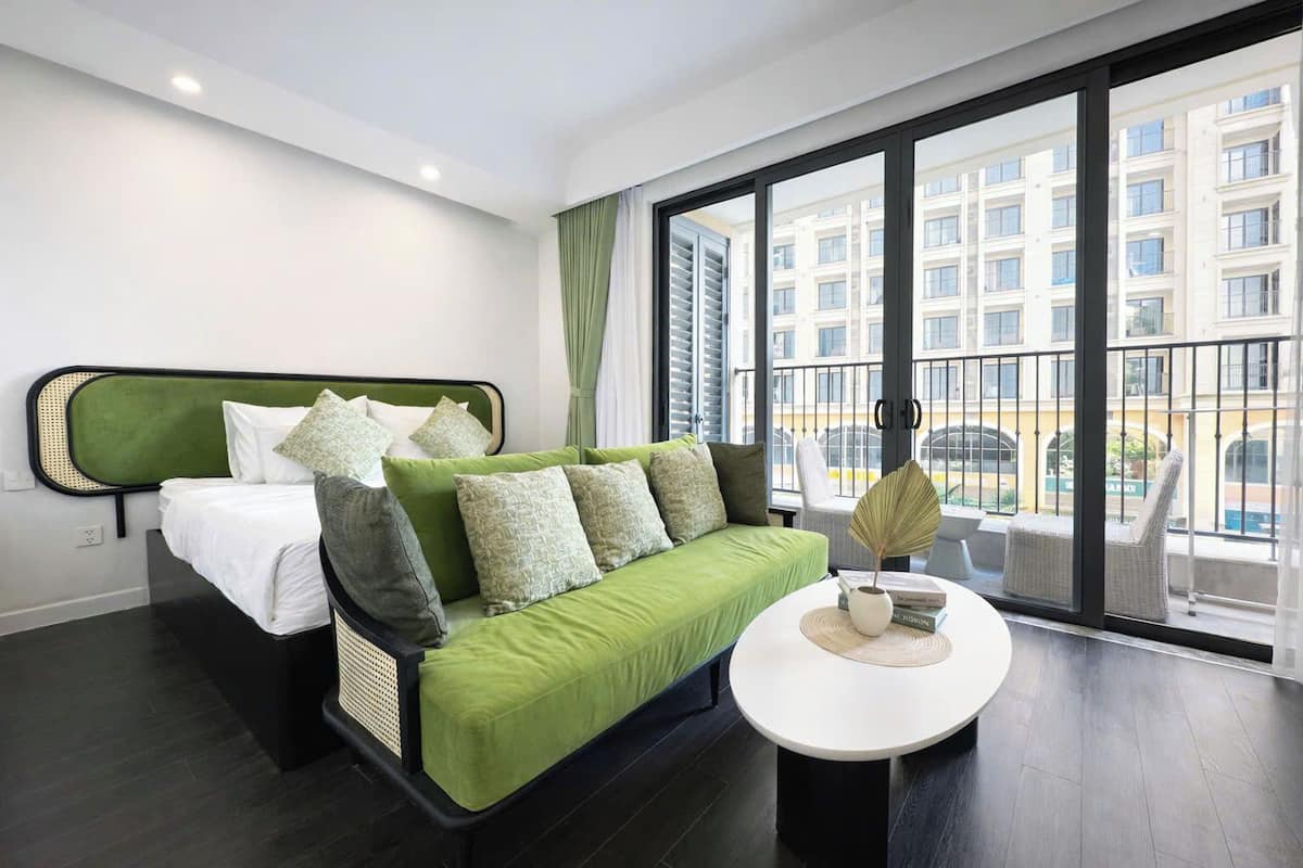 Quarto de hotel moderno com um sofá verde, uma mesa redonda branca e grandes janelas com vista para a paisagem urbana.