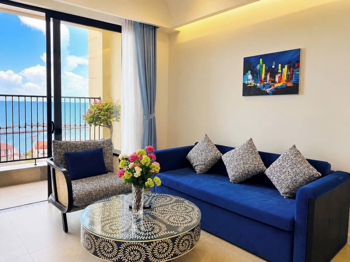 Varanda com vista para o mar, sofá azul com almofadas estampadas, mesa de centro de vidro com um vaso de flores e um quadro na parede.
