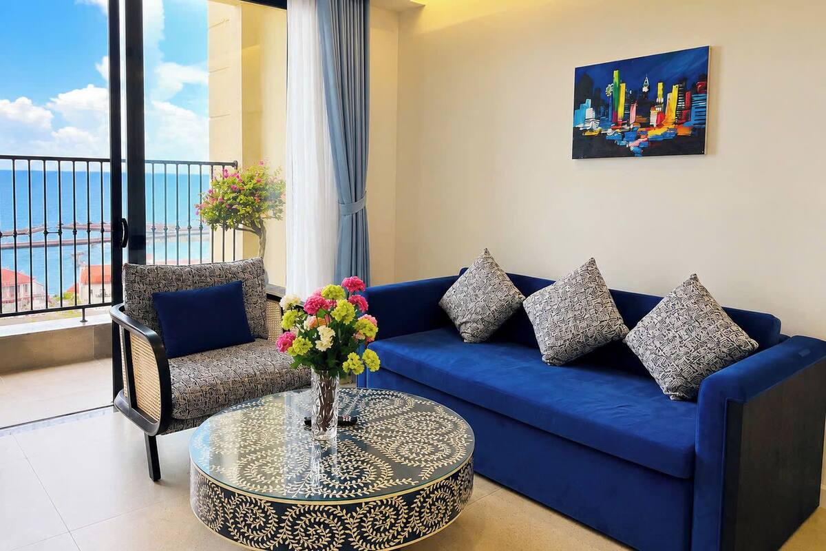 Varanda com vista para o mar, sofá azul com almofadas estampadas, mesa de centro de vidro com um vaso de flores e um quadro na parede.