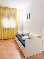 Acomodação pequena, com um quarto único, uma cama, uma janela com cortinas amarelas e um ponto de luz fixado na parede.