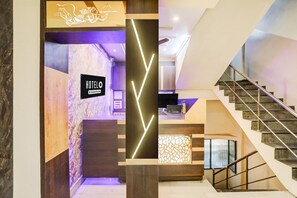 Uma área de recepção de hotel com design moderno, incluindo um balcão de atendimento, uma escadaria e uma placa com o nome do hotel.