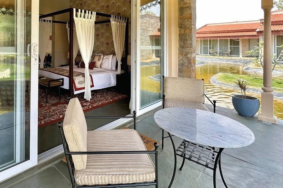Quarto com cama de dossel, área de estar com mesa e cadeiras, vista para piscina e edifício.