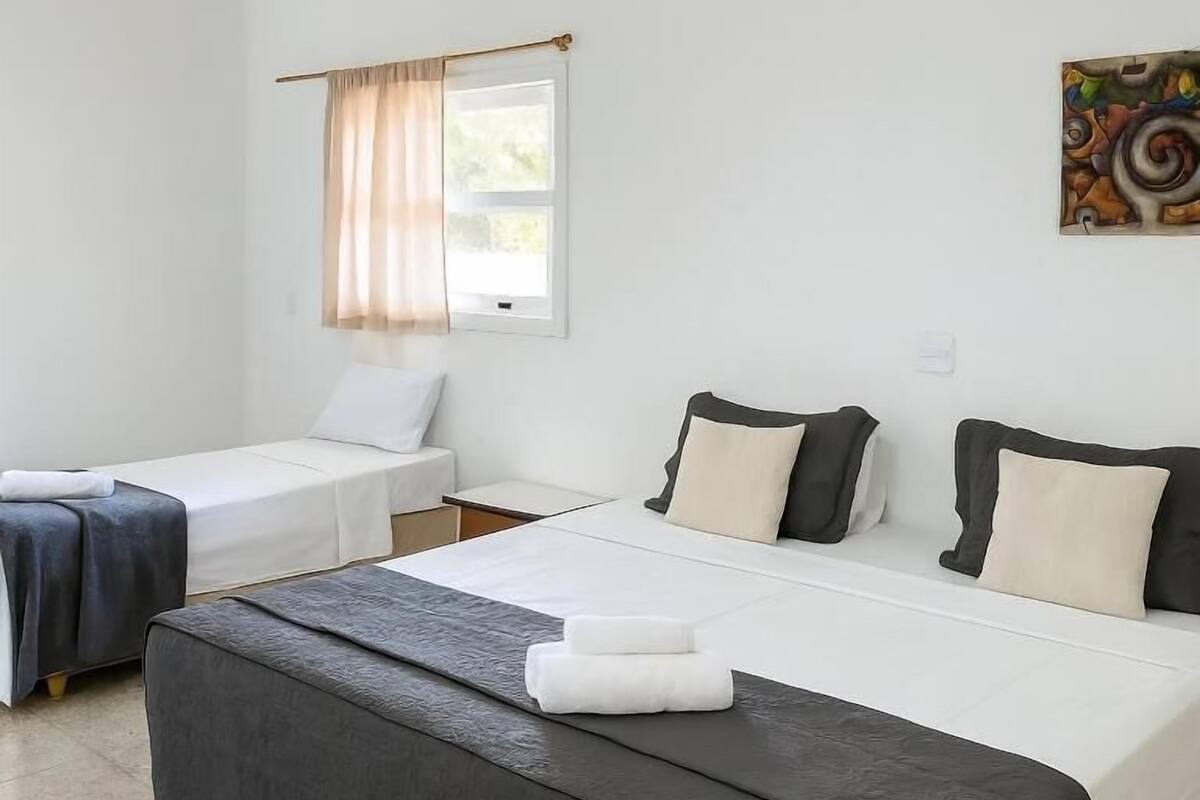 Quarto de hotel com duas camas de solteiro, uma cama de casal, uma janela com cortinas e um quadro na parede.
