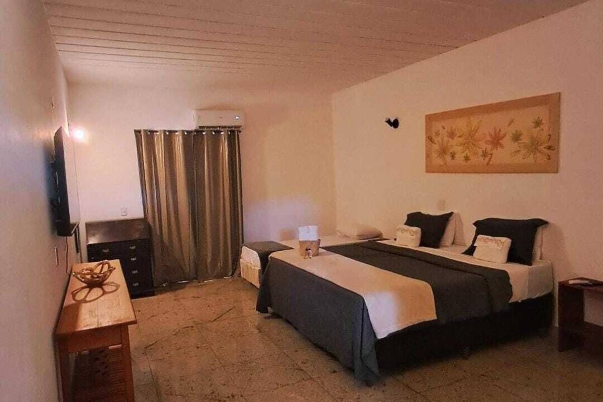 Quarto com cama, escrivaninha, cadeira, janela com cortinas e ar-condicionado fixado na parede.