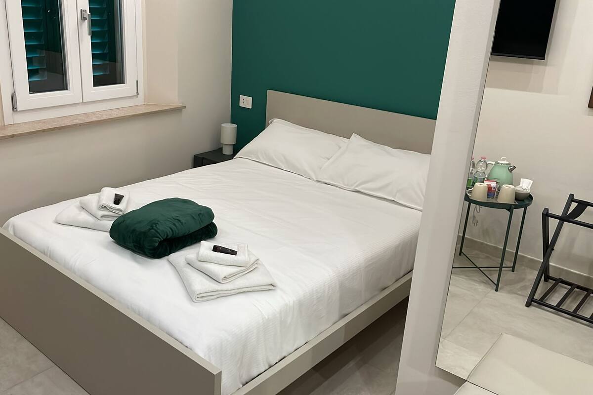 Cama bem arrumada com almofadas e toalhas dobradas, mesa de cabeceira com abajur, e janela com persianas.