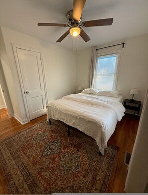 Quarto com cama, ventilador de teto, janela com persianas e tapete.
