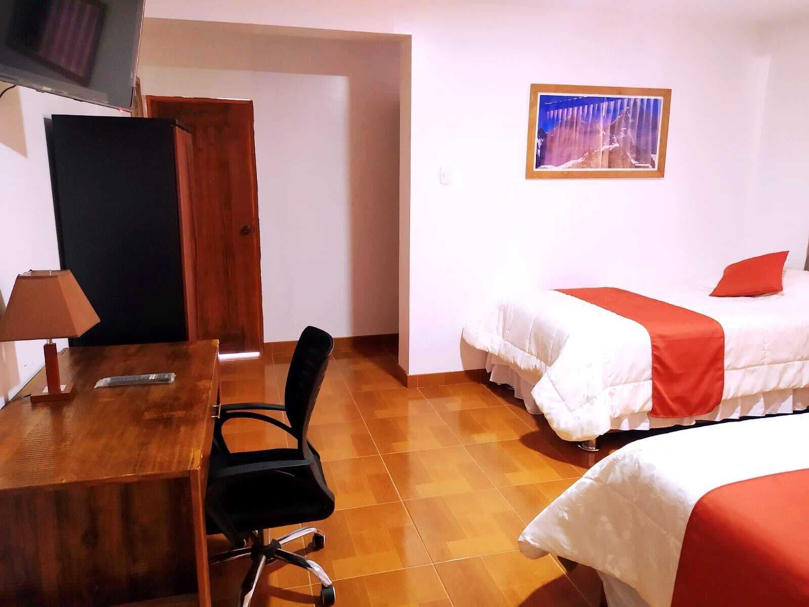 Quarto de hotel com escrivaninha de madeira, uma cadeira, duas camas com almofadas vermelhas, um quadro na parede e uma televisão à esquerda.