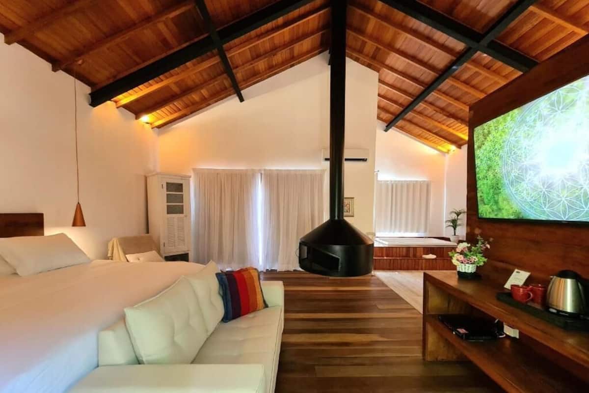 Um quarto amplo com teto alto, uma cama, um sofá, piso de madeira, lareira e televisão.