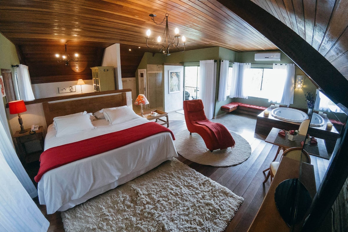 Quarto com uma cama grande, uma cadeira vermelha, uma mesa redonda com velas e vista para o exterior.