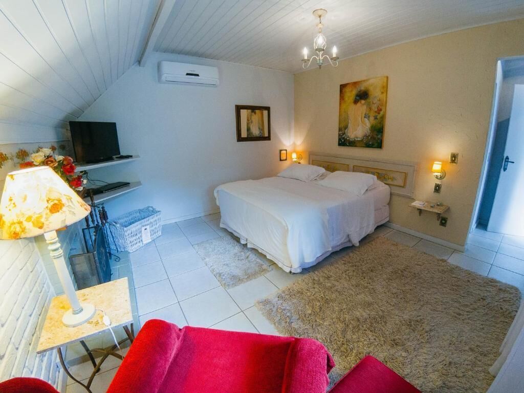 Quarto com cama, televisão, escrivaninha com abajur, uma cadeira vermelha e um quadro na parede.