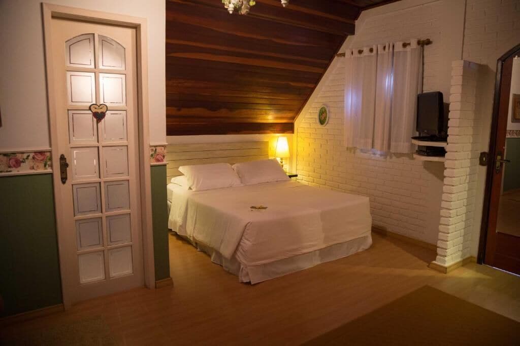 Quarto com cama, porta decorada com um coração, parede de tijolos, janela com cortinas e televisão em uma estante de tijolos.