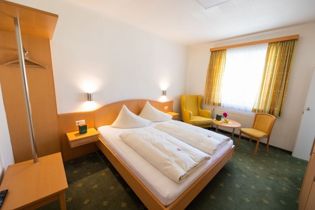 Quarto de hotel com cama, mesas de cabeceira, uma cadeira, uma pequena mesa e uma janela com cortinas.