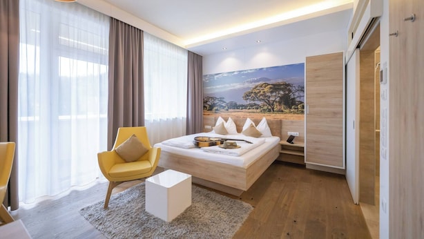 Quarto de hotel moderno com uma cama grande, uma poltrona amarela, uma mesa pequena branca e um quadro de uma paisagem na parede.