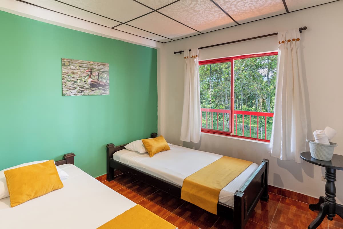 Quarto com duas camas, janela com grades vermelhas, parede verde e um quadro.