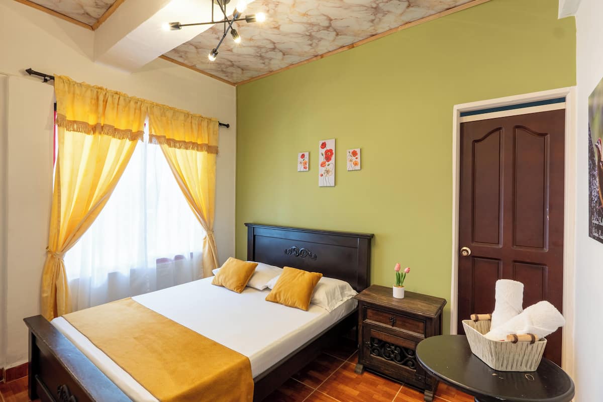 Um quarto com cama, cortinas, mesa de cabeceira, uma pequena mesa e uma porta.
