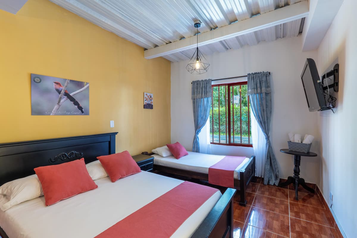Quarto com duas camas, um ventilador de teto, janela com cortinas e televisão presa na parede.