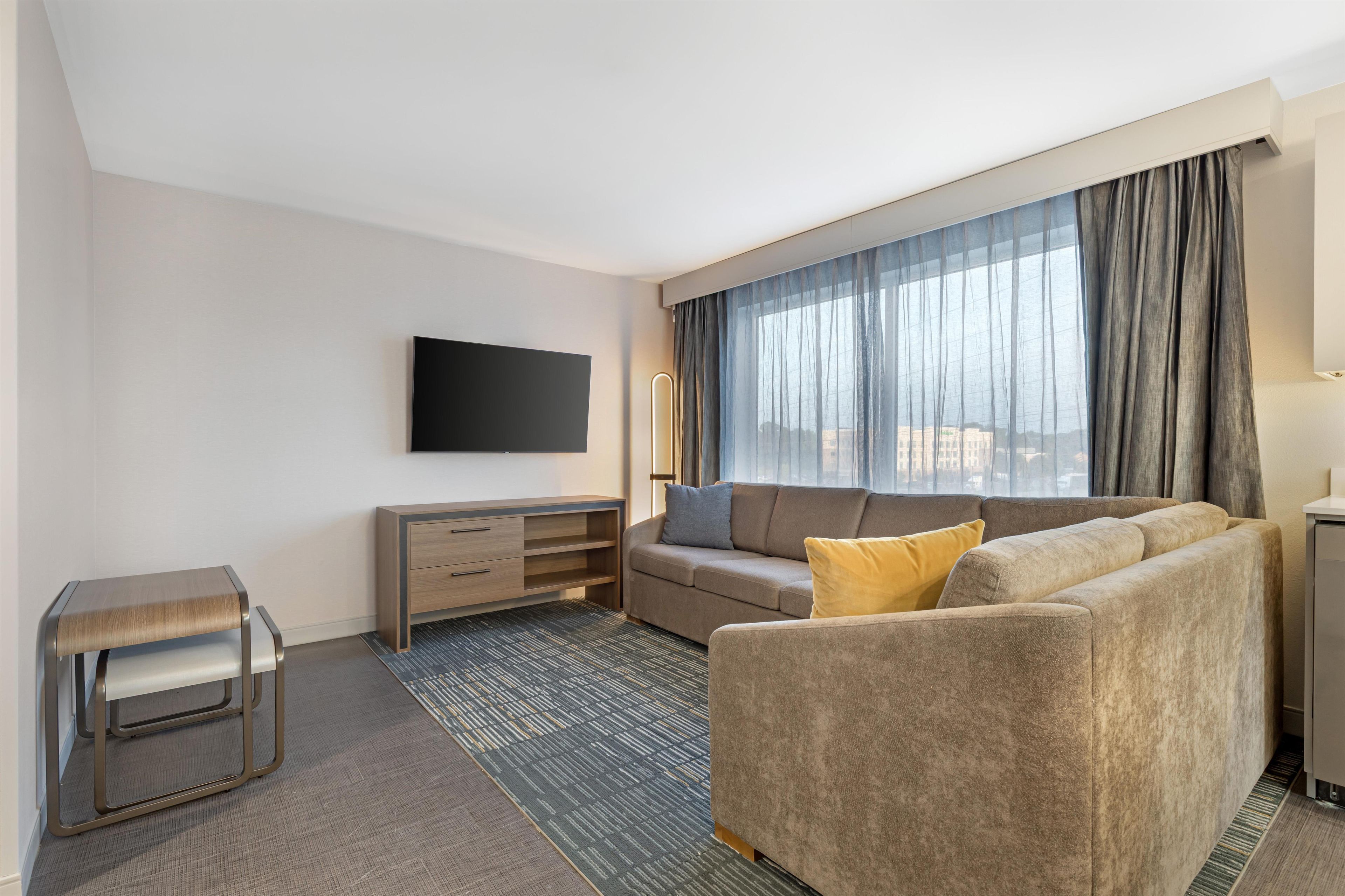 Quarto de hotel moderno com sofá, TV de tela plana fixada na parede, mesa de cabeceira e janela com cortinas.