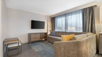 Quarto de hotel moderno com sofá, TV de tela plana fixada na parede, mesa de cabeceira e janela com cortinas.