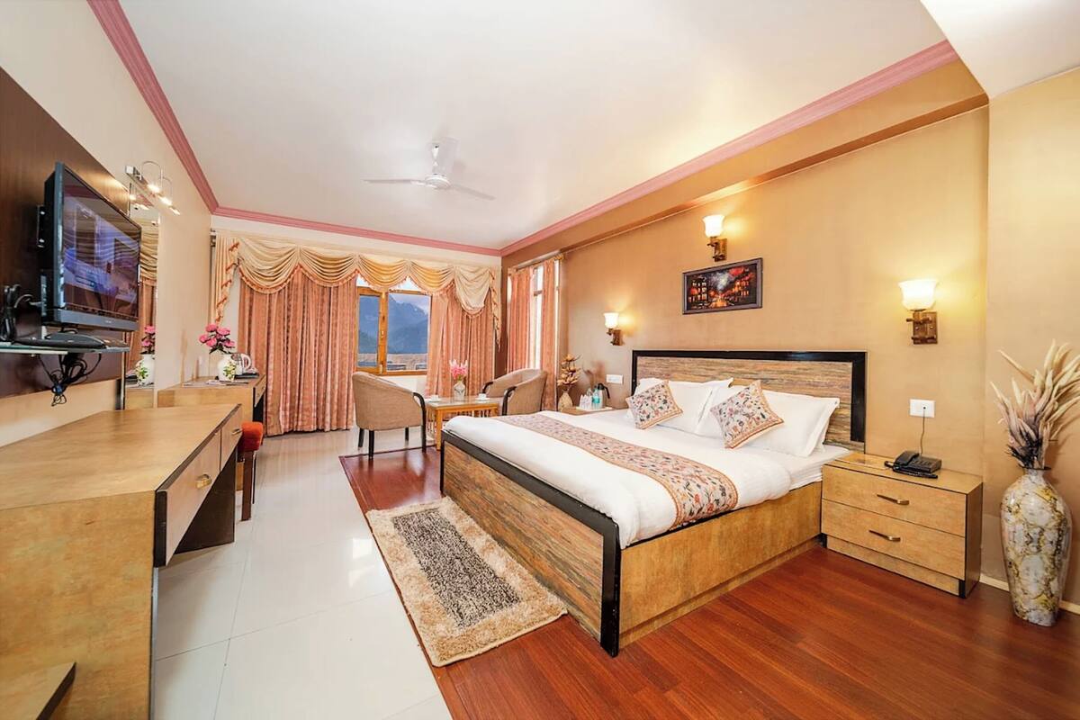 Quarto de hotel com cama, mesas de cabeceira, escrivaninha com televisão, cadeira, janela com cortinas e um vaso com plantas secas.