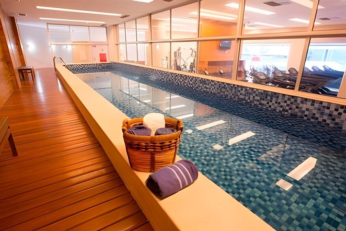Piscina interior comprida e estreita, com borda em azulejo, soalho de madeira e um cesto com toalhas e um travesseiro ao lado.