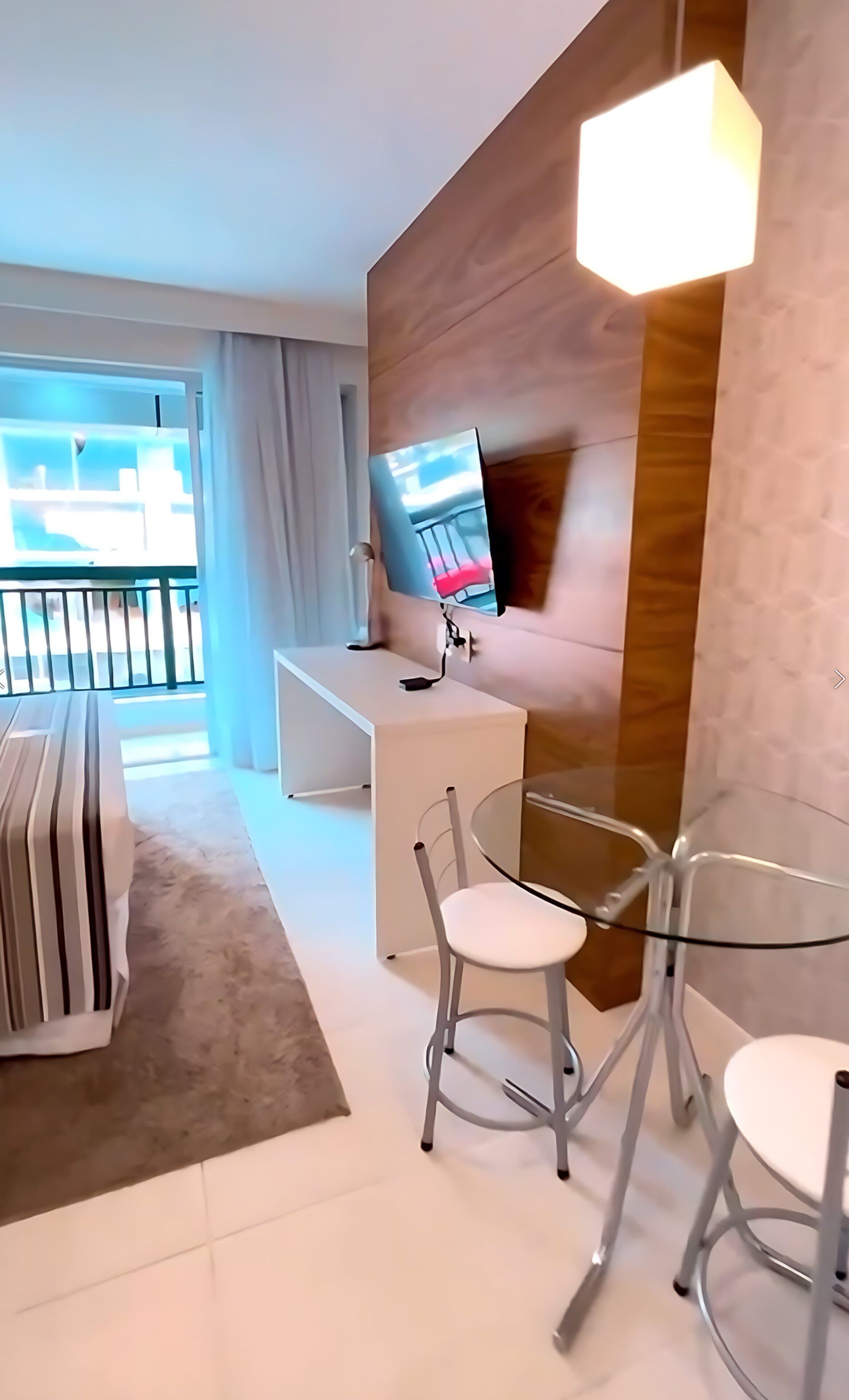 Quarto de hotel moderno com uma grande TV de tela plana, uma escrivaninha branca, dois banquetes de vidro, uma cama grande e uma varanda com vista.