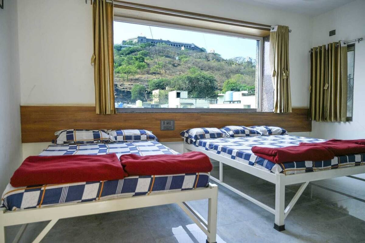 Quarto com duas camas, uma janela grande com cortinas e vista para uma colina com edifícios.
