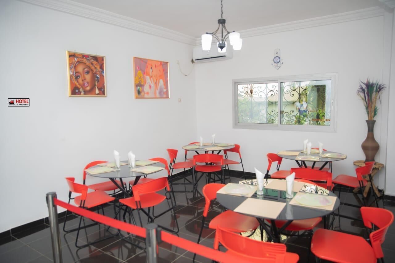 Restaurante com mesas redondas e cadeiras vermelhas, duas pinturas na parede, um candelabro e uma janela com detalhes em ferro.