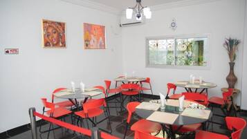 Restaurante com mesas redondas e cadeiras vermelhas, duas pinturas na parede, um candelabro e uma janela com detalhes em ferro.