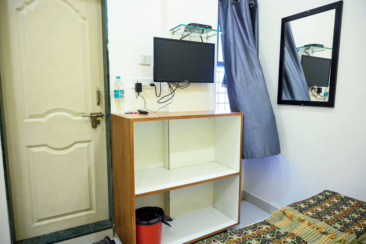 Quarto com prateleira de madeira, televisão na parede, espelho e porta.