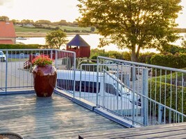 Um deck de madeira com guarda-corpo, um vaso de planta e vista para a água e edifícios ao longe.