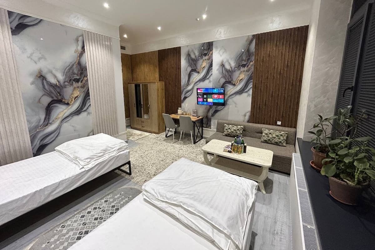 Quarto de hotel moderno com duas camas de solteiro, um sofá, uma mesinha, uma TV e um vaso de plantas.