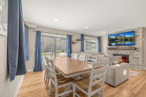 Uma sala de estar moderna com lareira, área de jantar com mesa de madeira e cadeiras brancas, e uma grande TV de tela plana fixada numa parede de pedra.