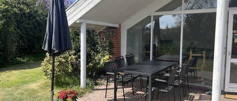 Um alpendre coberto com mesa, cadeiras, um vaso de planta e um guarda-sol.