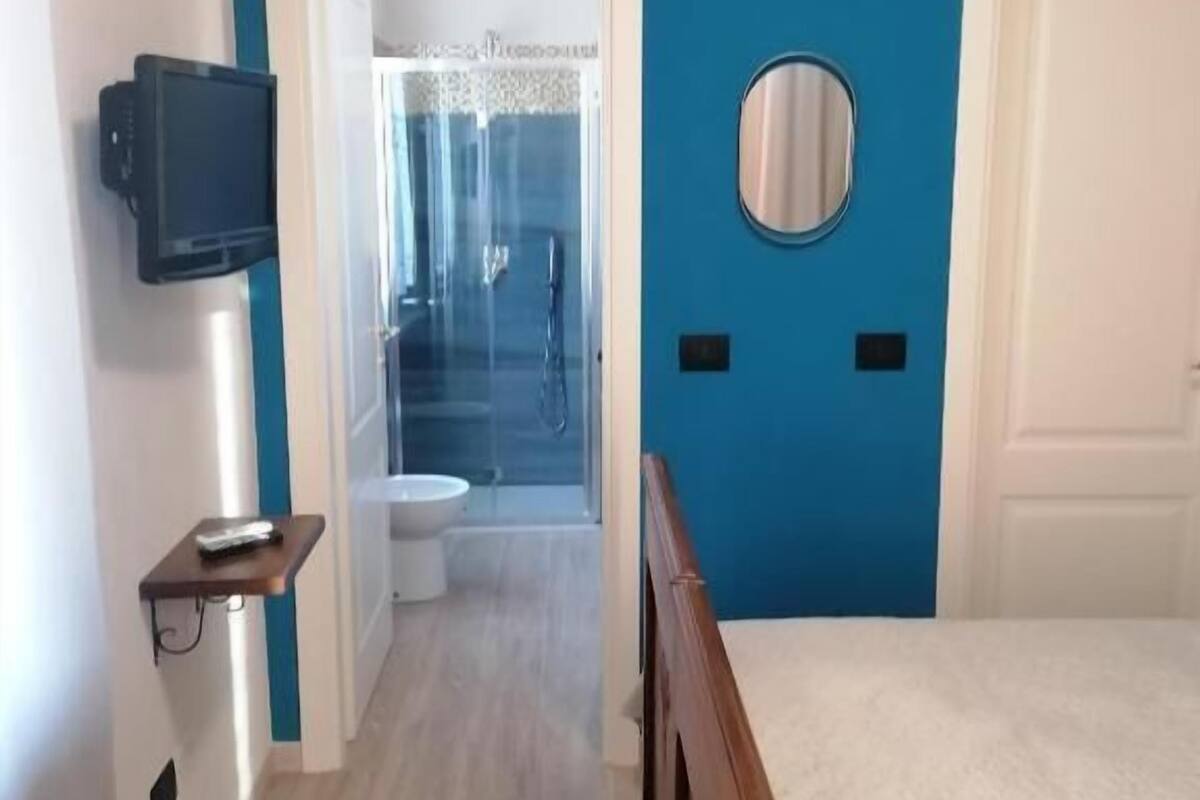 Quarto com cama, televisão fixada na parede, espelho e banheiro visível através de uma porta aberta.