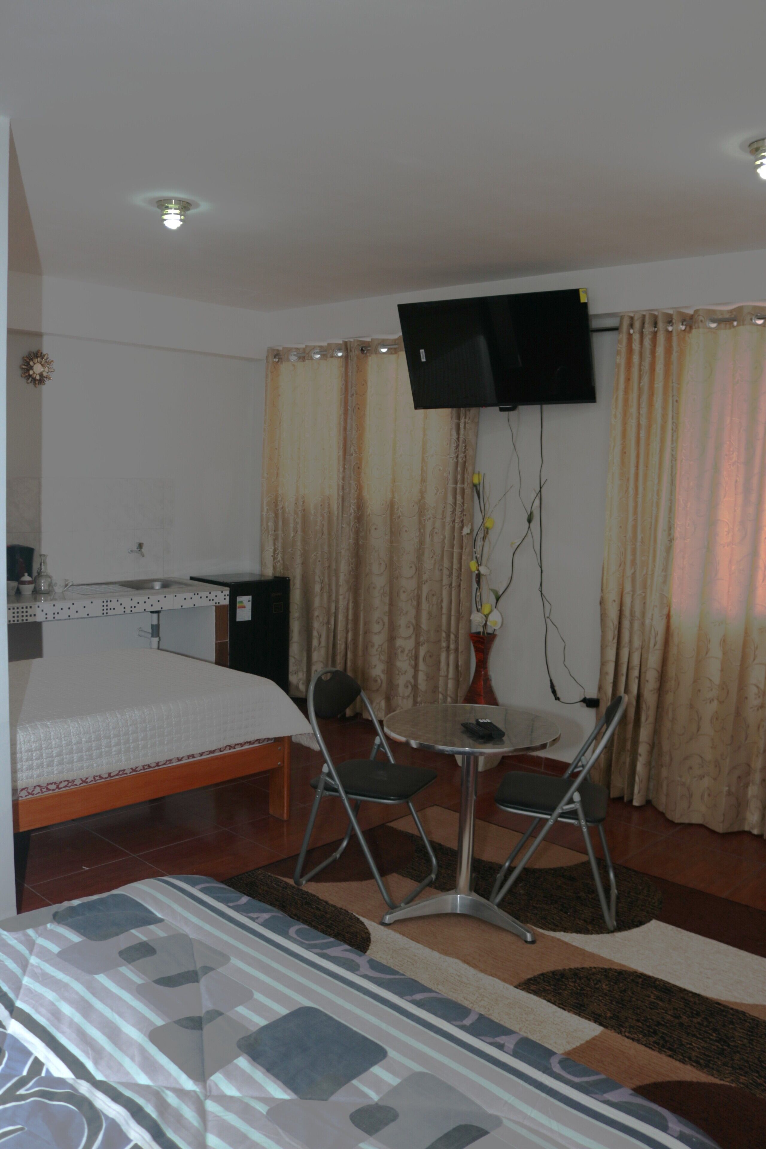 Quarto de hotel com cama, escrivaninha, cadeira, TV fixada na parede e cortinas.