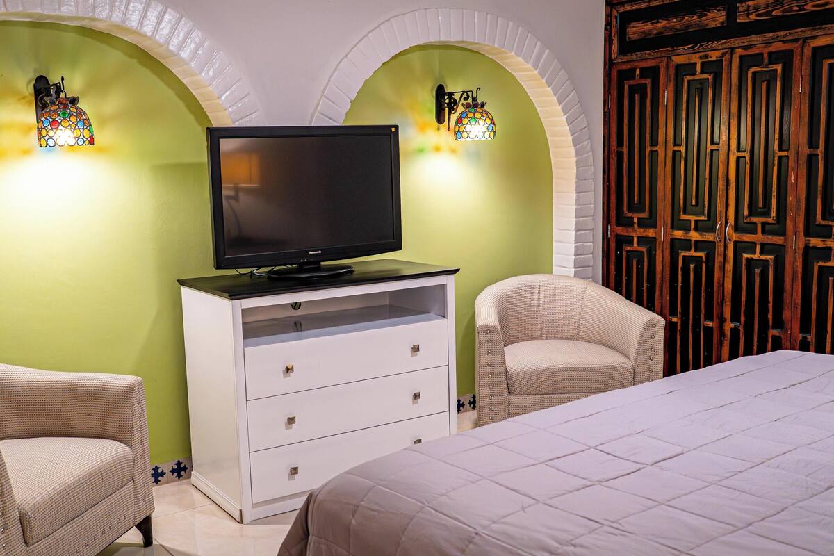 Quarto com televisão sobre um armário branco, duas cadeiras brancas, uma cama e uma porta de madeira com padrão geométrico.